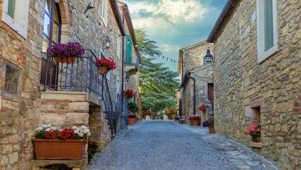 Uma rua de paralelepípedos estreita, ladeada por edifícios de pedra com caixas de flores e luminárias pendentes.
