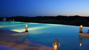 Uma piscina infinita com duas luzes em estilo de lanterna nos cantos, cercada por uma área ajardinada com vista para colinas ao longe.