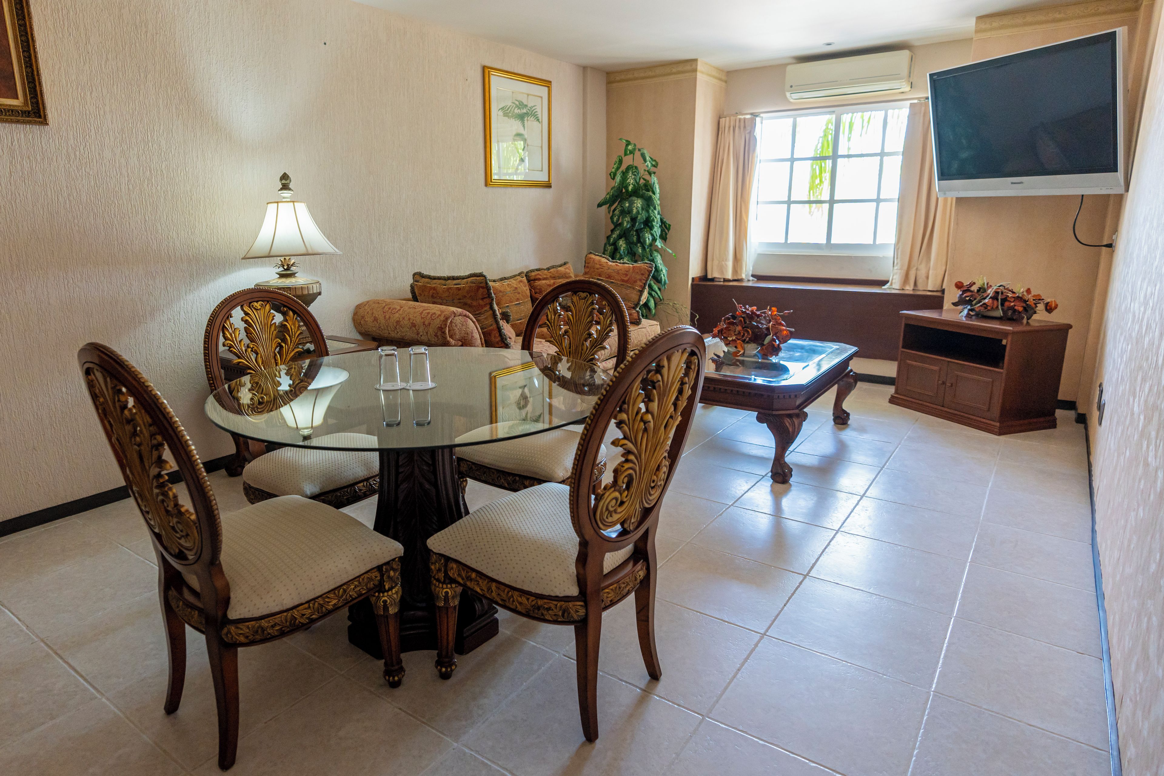 Uma sala de jantar com mesa de vidro rodeada por cadeiras elaboradas, um sofá com almofadas estampadas, um abajur decorativo e uma televisão na parede.