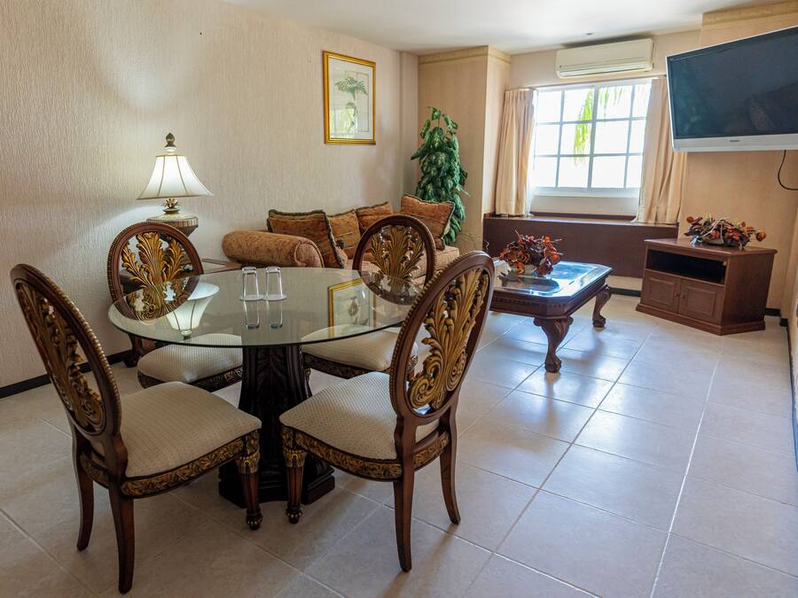 Uma sala de jantar com mesa de vidro rodeada por cadeiras elaboradas, um sofá com almofadas estampadas, um abajur decorativo e uma televisão na parede.