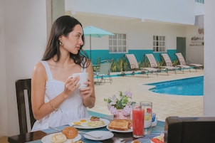 Uma pessoa sentada à mesa com itens de café da manhã, uma piscina e cadeiras de sol ao fundo.
