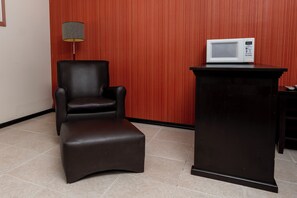 Quarto com poltrona de couro marrom, um pequeno banquinho, armário de madeira escura e micro-ondas branca em cima.