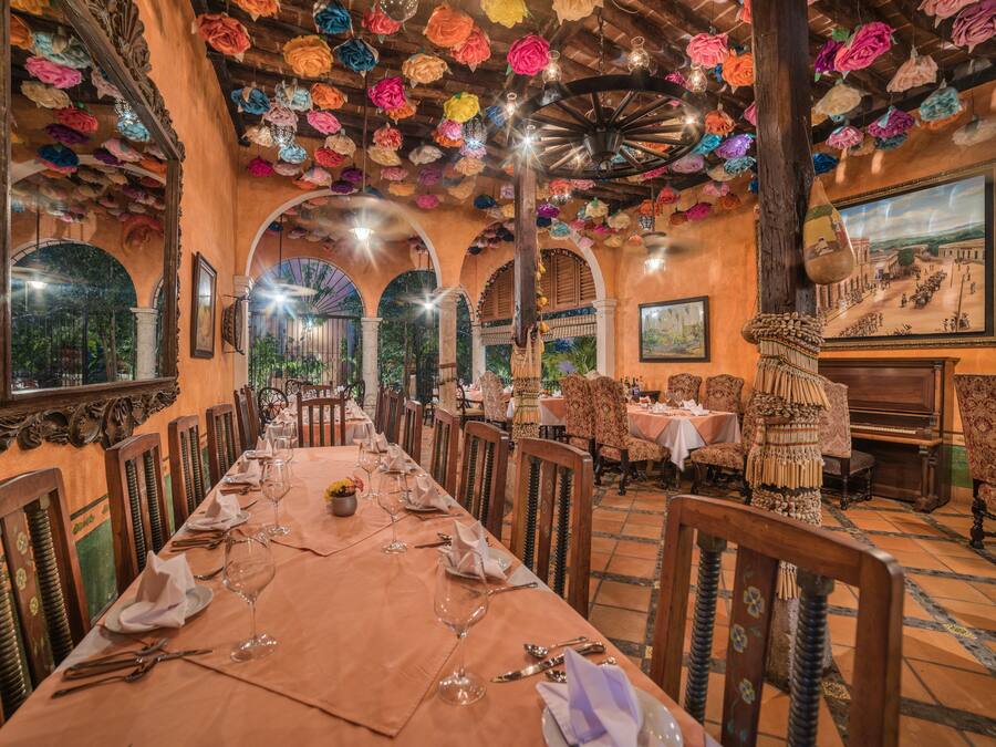 Uma longa mesa de jantar posta com cadeiras, decoração de teto ornamentada e um piano em um ambiente bem iluminado com arcos e um mural.