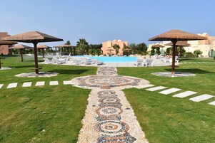 Área da piscina com espreguiçadeiras e guarda-sóis, um caminho com padrão e um edifício ao fundo.