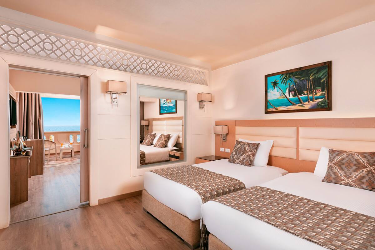 Quarto de hotel com duas camas, uma janela grande, um quadro com uma cena de praia e vista para o mar.
