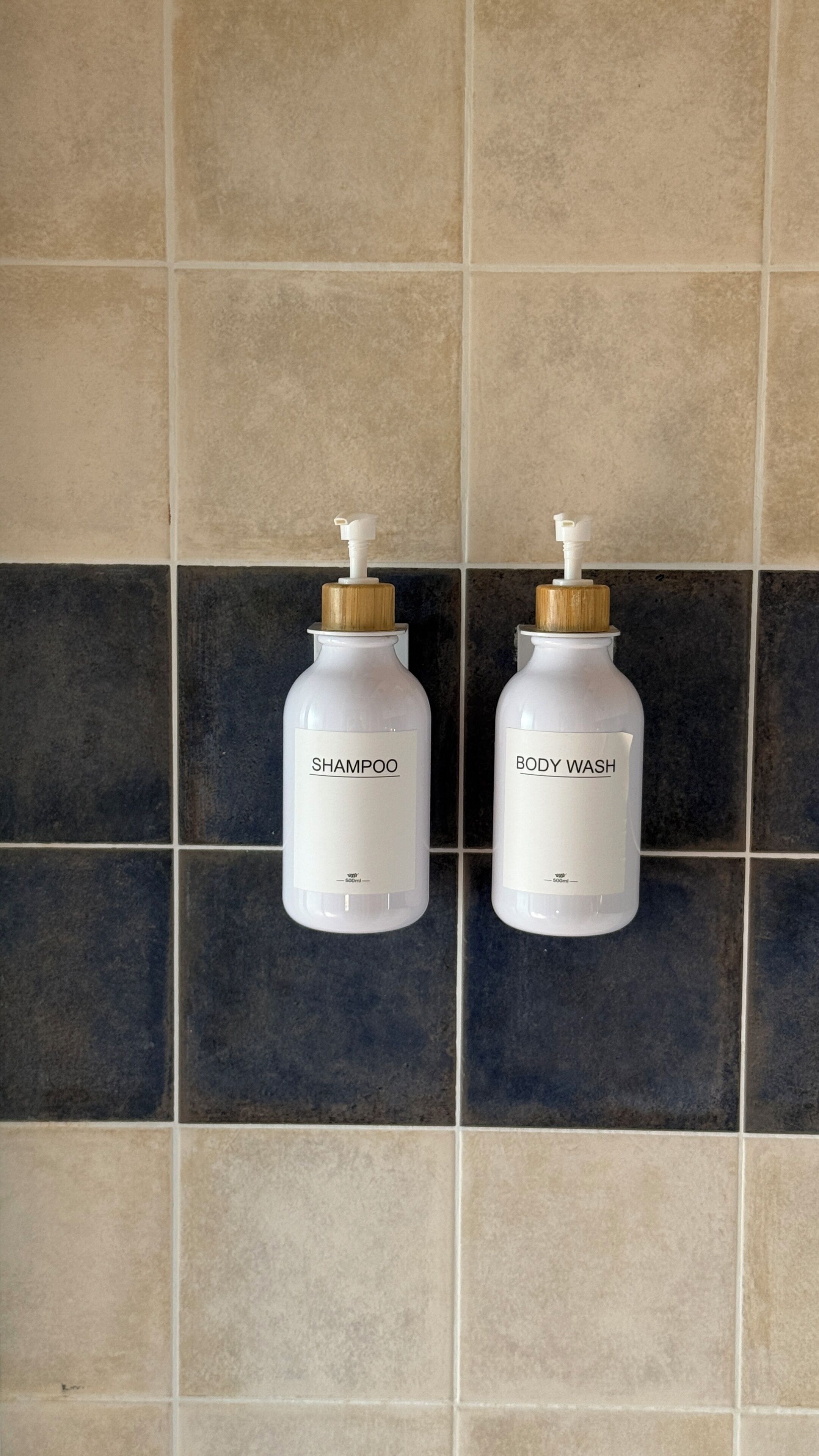 Duas garrafas brancas com bombas, rotuladas 'SHAMPOO' e 'BODY WASH', fixadas numa parede revestida a azulejos.