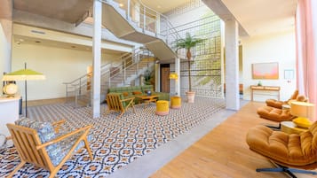 Um hall amplo com piso estampado, uma escada, um sofá verde, uma cadeira de madeira e um banquinho amarelo.