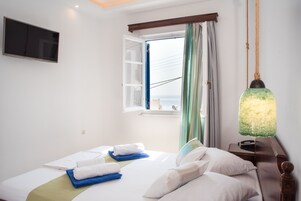 Quarto de hotel com uma cama, televisão na parede, janela com cortinas e mesa de cabeceira com abajur.