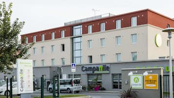 Um hotel com fachada moderna, uma placa com o nome Campanile Hotel Restaurant, e um ônibus indicando acesso por transporte público.