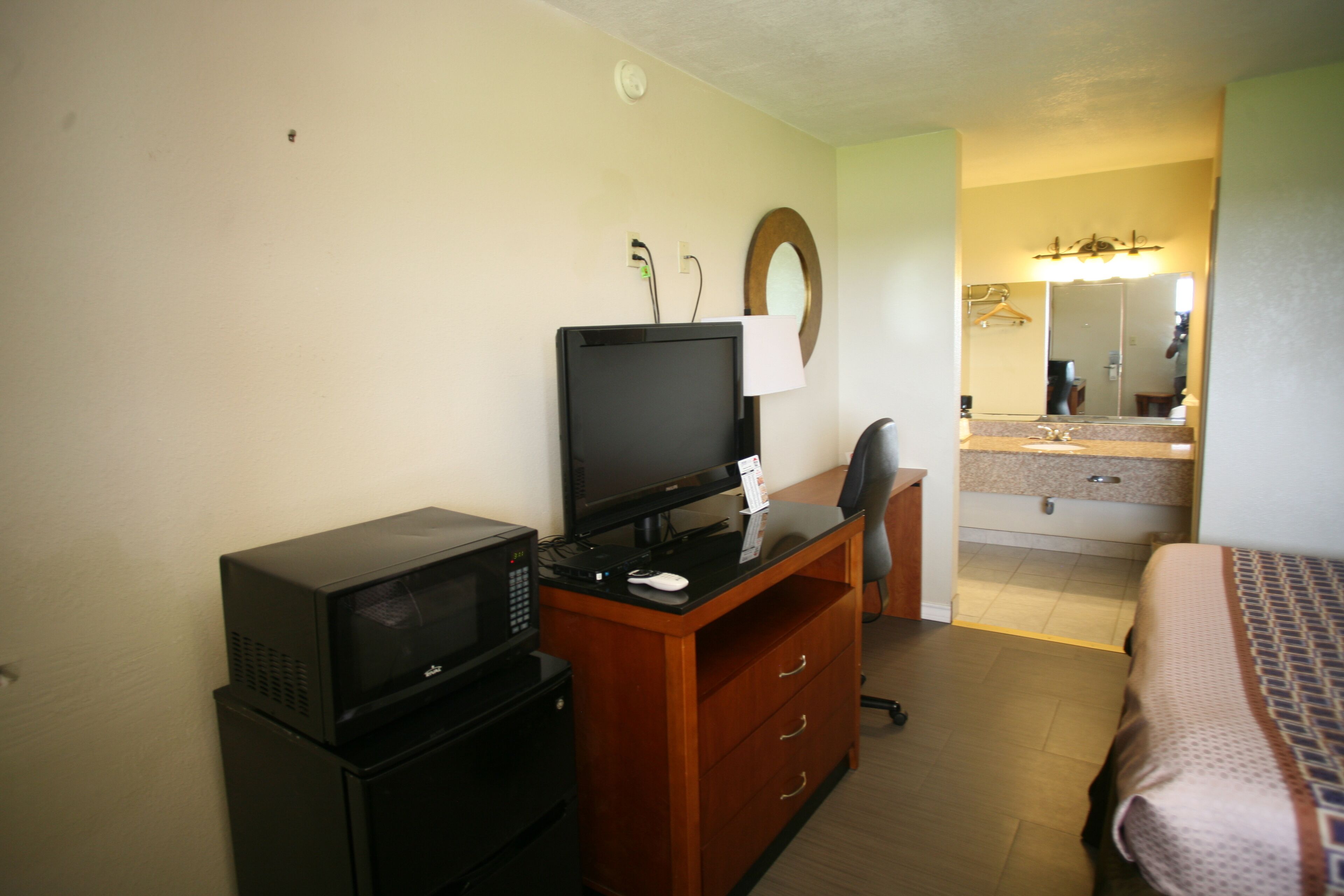 Quarto de hotel com escrivaninha, televisão, micro-ondas, cama, penteadeira, espelho e banheiro visível através de uma porta aberta.