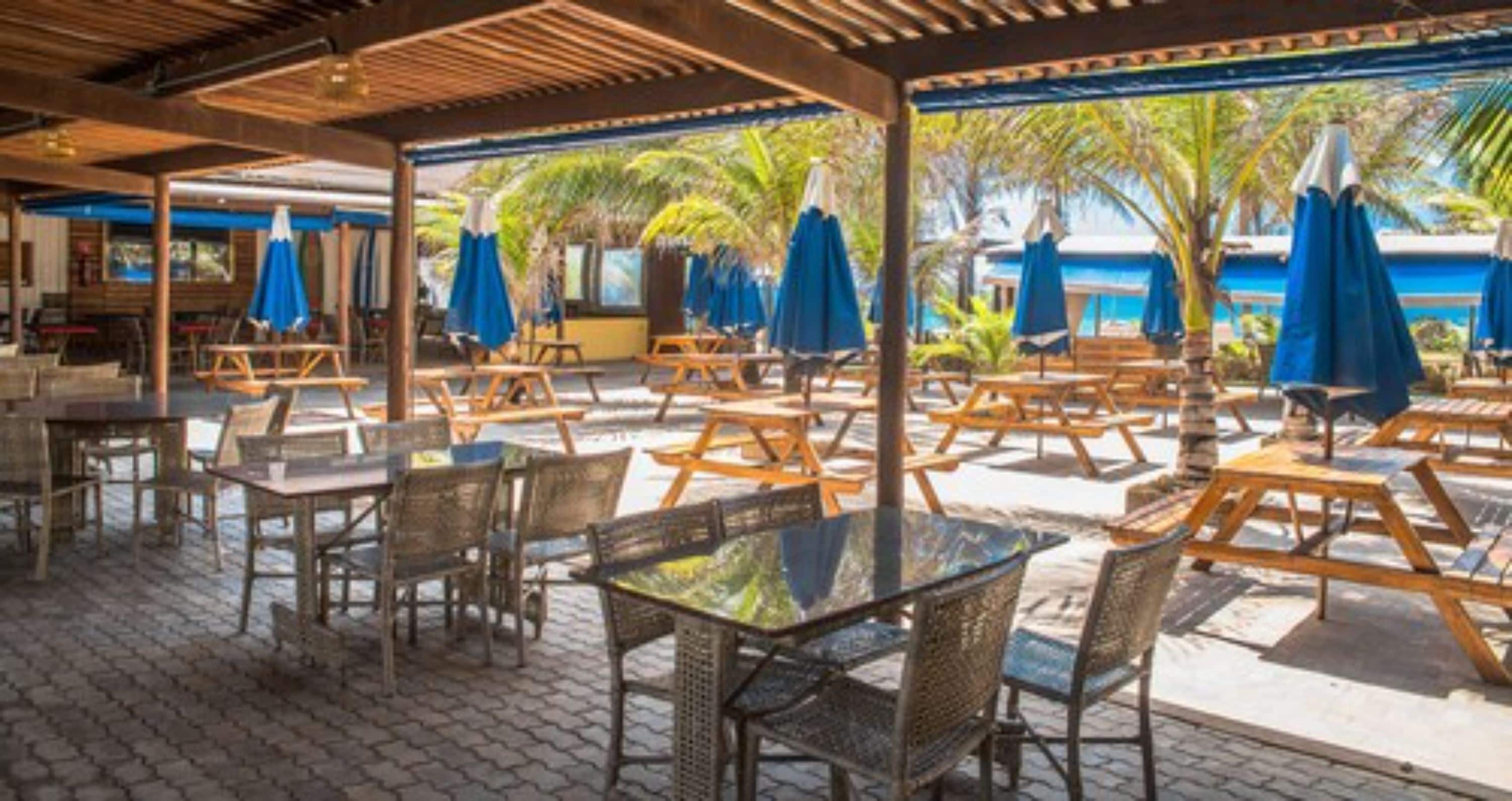 Um café ao ar livre com mesas e cadeiras de madeira, guarda-sóis azuis e piso de azulejos.