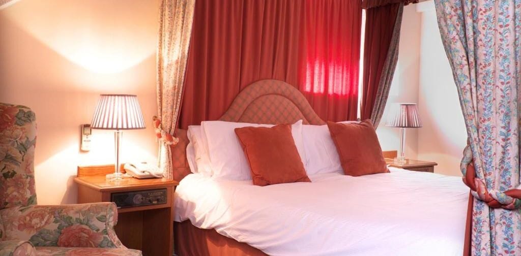 Quarto de hotel com uma cama, duas mesas de cabeceira, uma luminária e cortinas.