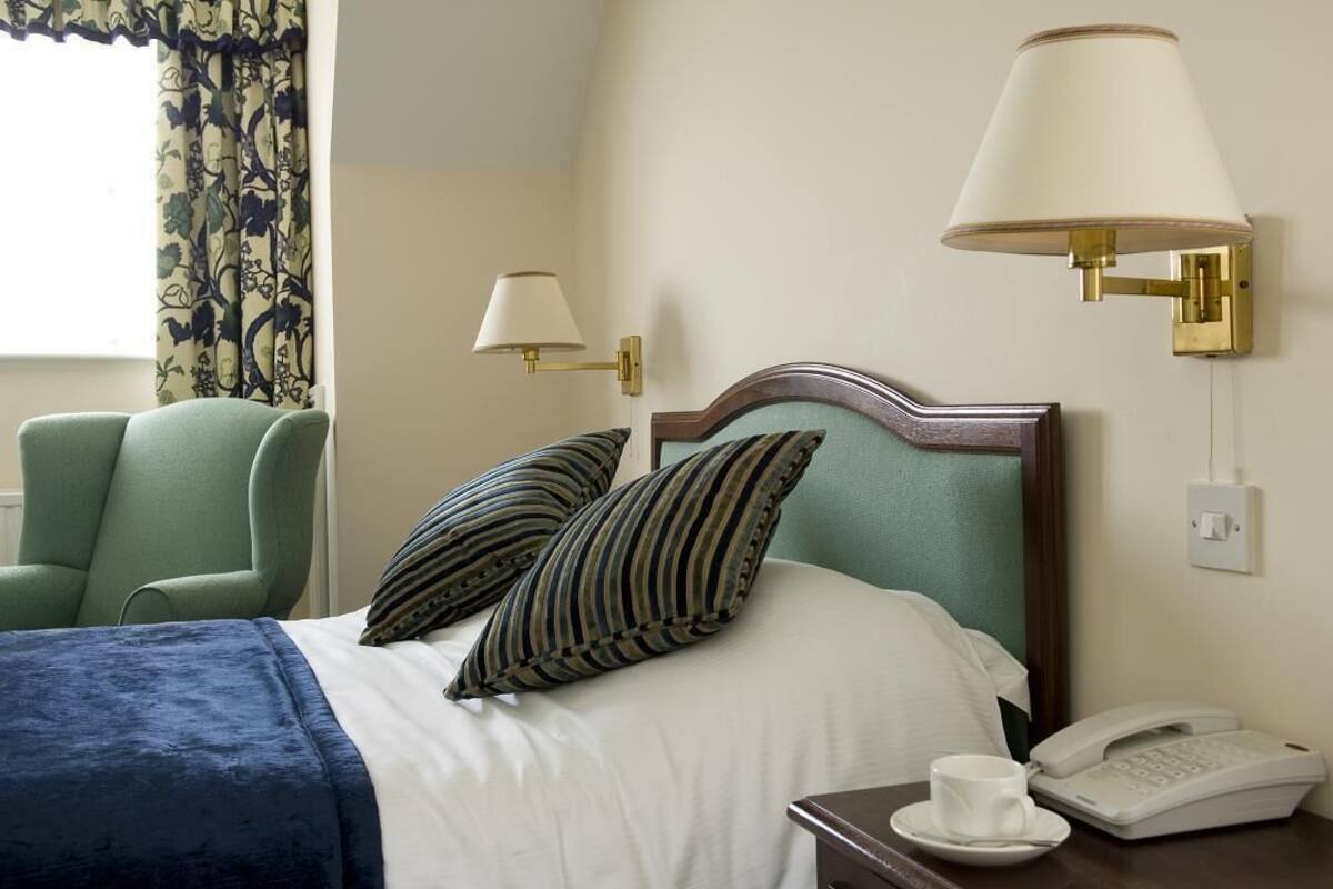 Quarto de hotel com cama, duas almofadas, mesa de cabeceira com telefone e chávena, cadeira e duas luminárias de parede.