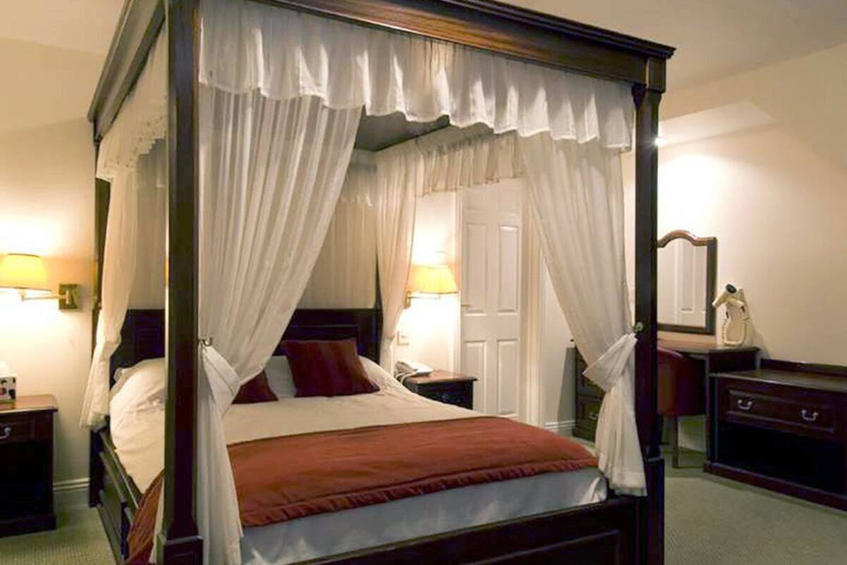 Uma cama de dossel com cortinas brancas, uma cabeceira de madeira e duas luminárias de cabeceira.