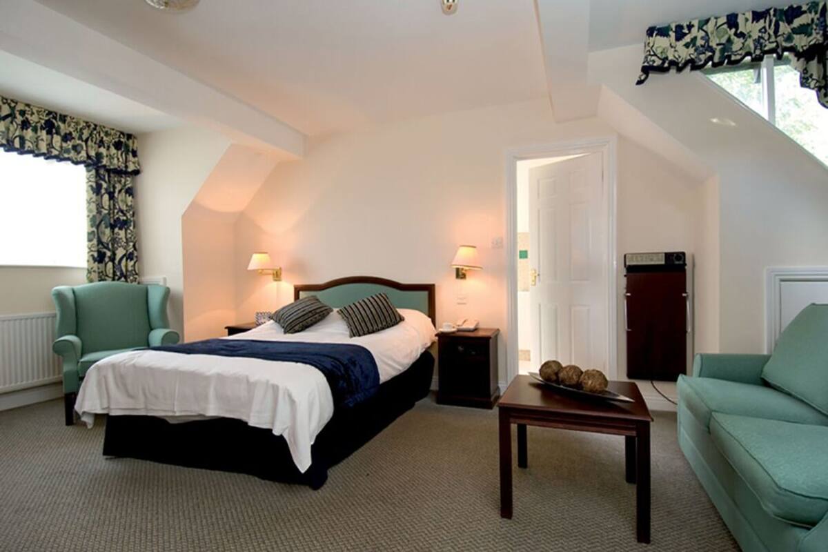 Quarto de hotel com cama, duas mesas de cabeceira, um criado-mudo, uma poltrona verde, um sofá verde e uma janela com cortinas.