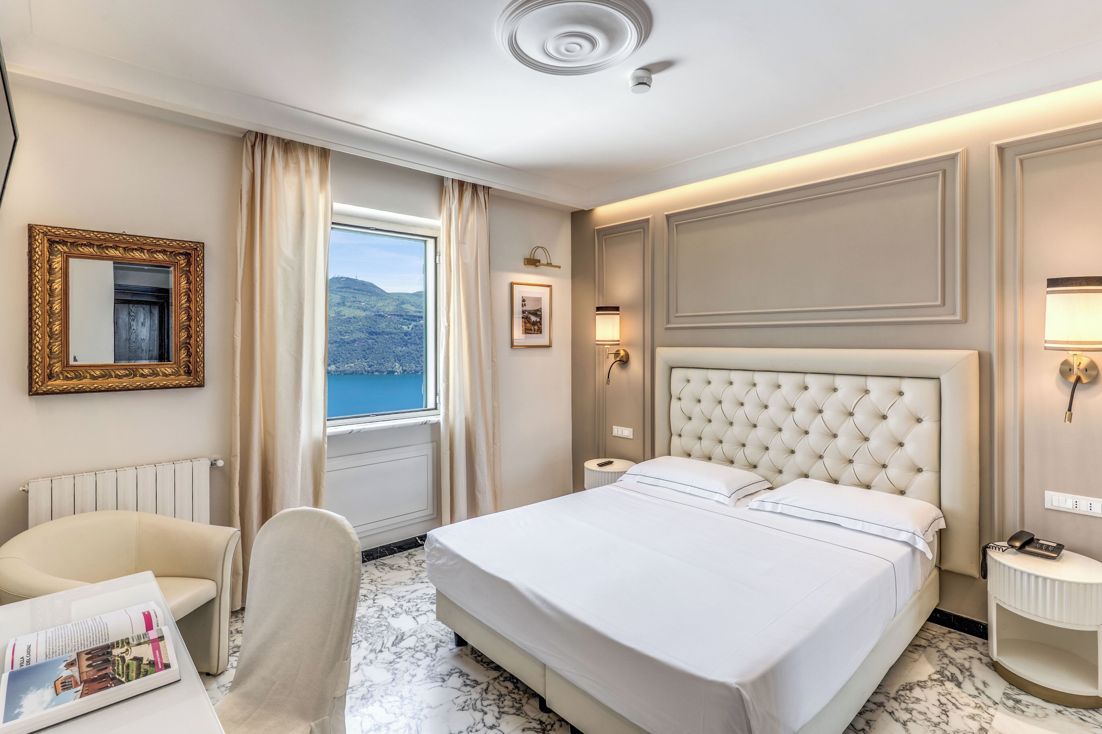 Quarto de hotel com uma cama grande, uma cadeira, uma mesa com uma revista, uma janela com vista e um espelho.