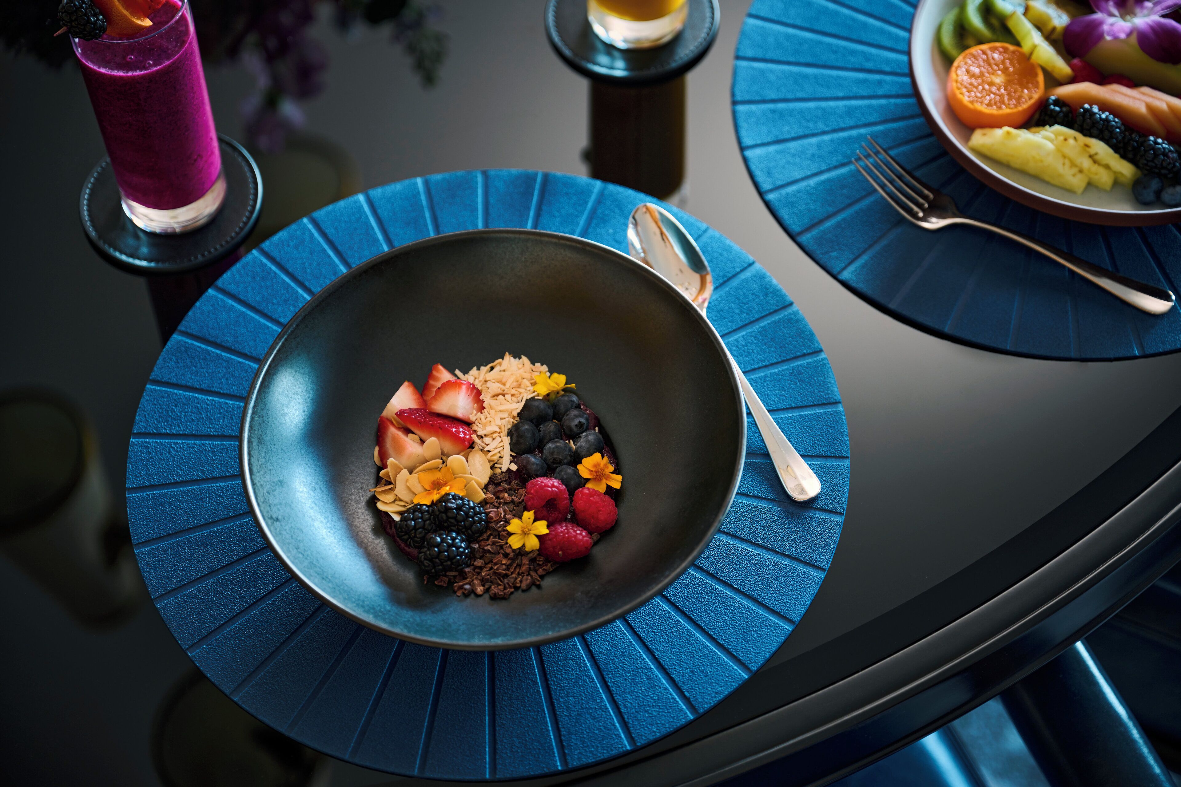 Uma mesa com um jogo americano azul, uma tigela de frutas vermelhas e nozes, um garfo e um copo de bebida roxa.