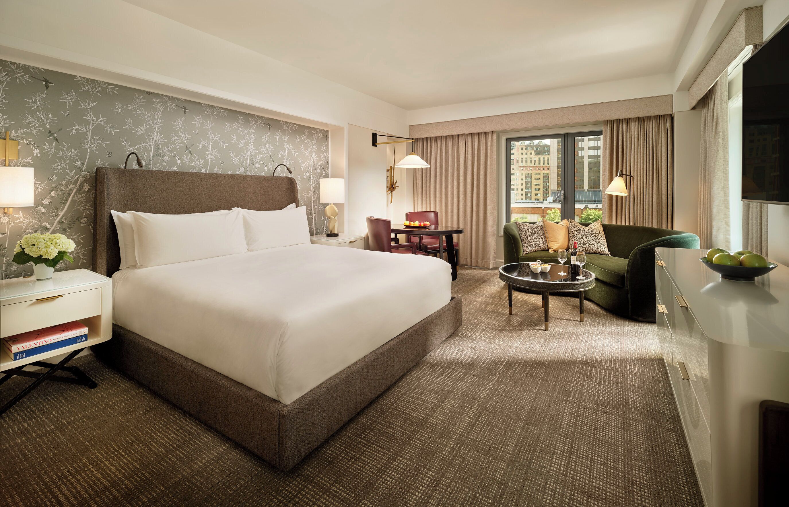 Quarto de hotel com uma cama grande, uma escrivaninha com abajur, uma cadeira, uma mesinha com uma cesta de frutas e uma televisão.