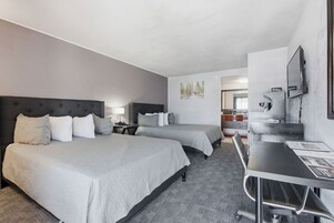 Quarto de hotel com duas camas, uma escrivaninha, uma cadeira, uma televisão e um banheiro ao fundo.
