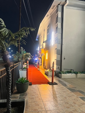 Vista noturna de uma rua com um tapete vermelho que leva a um edifício de fachada branca e várias janelas.