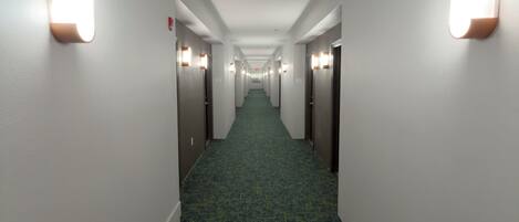 Corredor de hotel com piso carpete, paredes brancas e luminárias fixadas na parede.