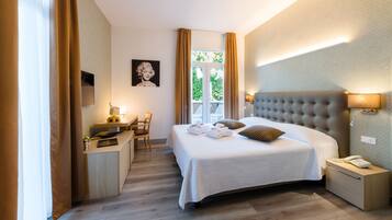 Quarto de hotel com cama, mesa de cabeceira, abajur e escrivaninha com cadeira.