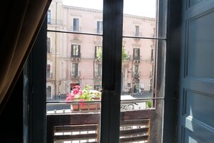 Vista da janela com varanda, mostrando um edifício de arquitetura clássica e uma rua.