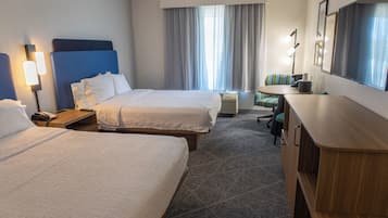 Quarto de hotel com duas camas, uma escrivaninha, uma cadeira, um abajur e uma janela com cortinas.