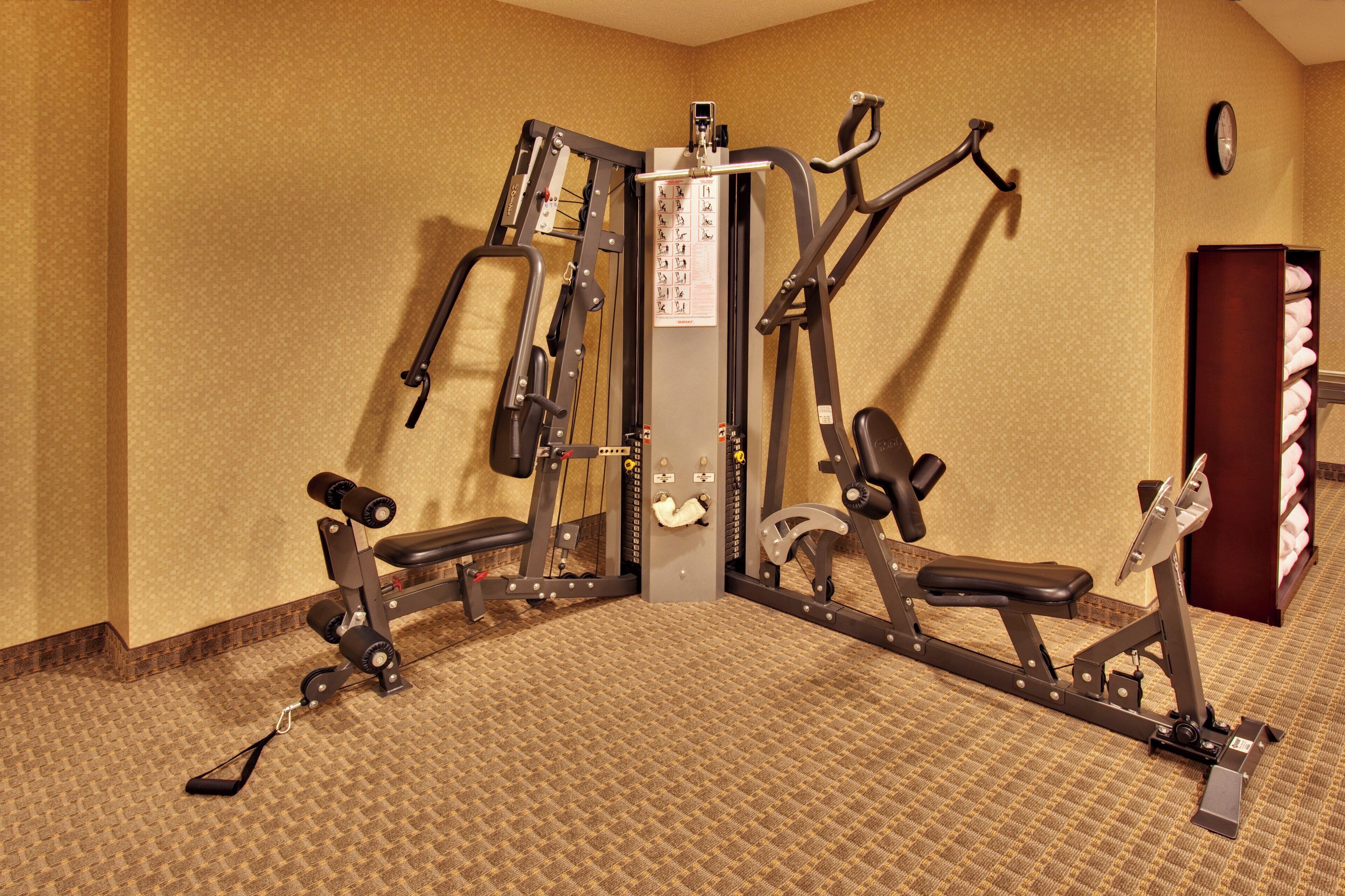 Quarto de hotel com área de ginásio, apresentando uma máquina de exercícios multifuncional, um relógio fixado na parede e um porta-toalhas.