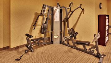 Quarto de hotel com área de ginásio, apresentando uma máquina de exercícios multifuncional, um relógio fixado na parede e um porta-toalhas.