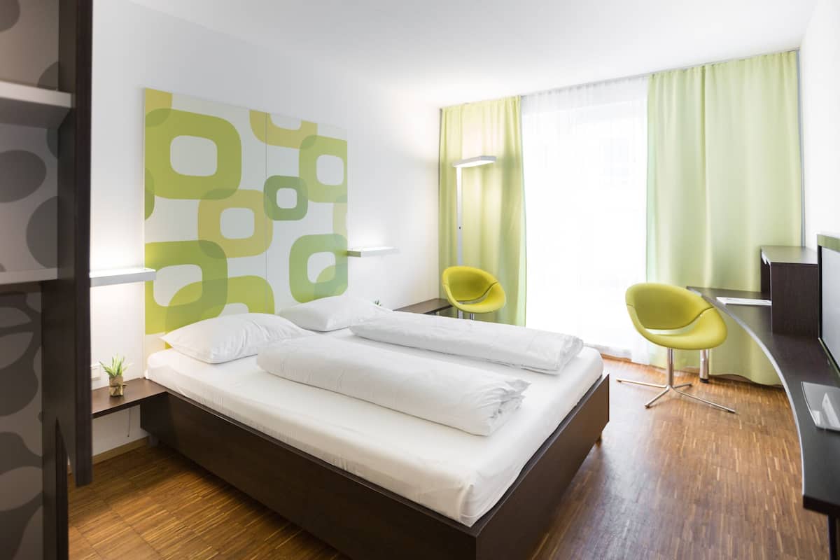 Quarto de hotel moderno com uma cama grande, uma mesa, uma cadeira e uma parede verde com padrões abstratos.