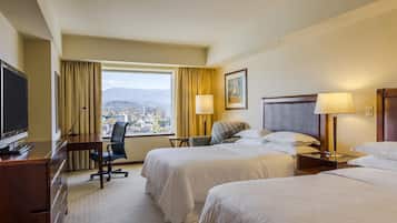 Quarto de hotel com duas camas, uma escrivaninha com televisão, uma cadeira, um abajur e vista para a paisagem urbana.