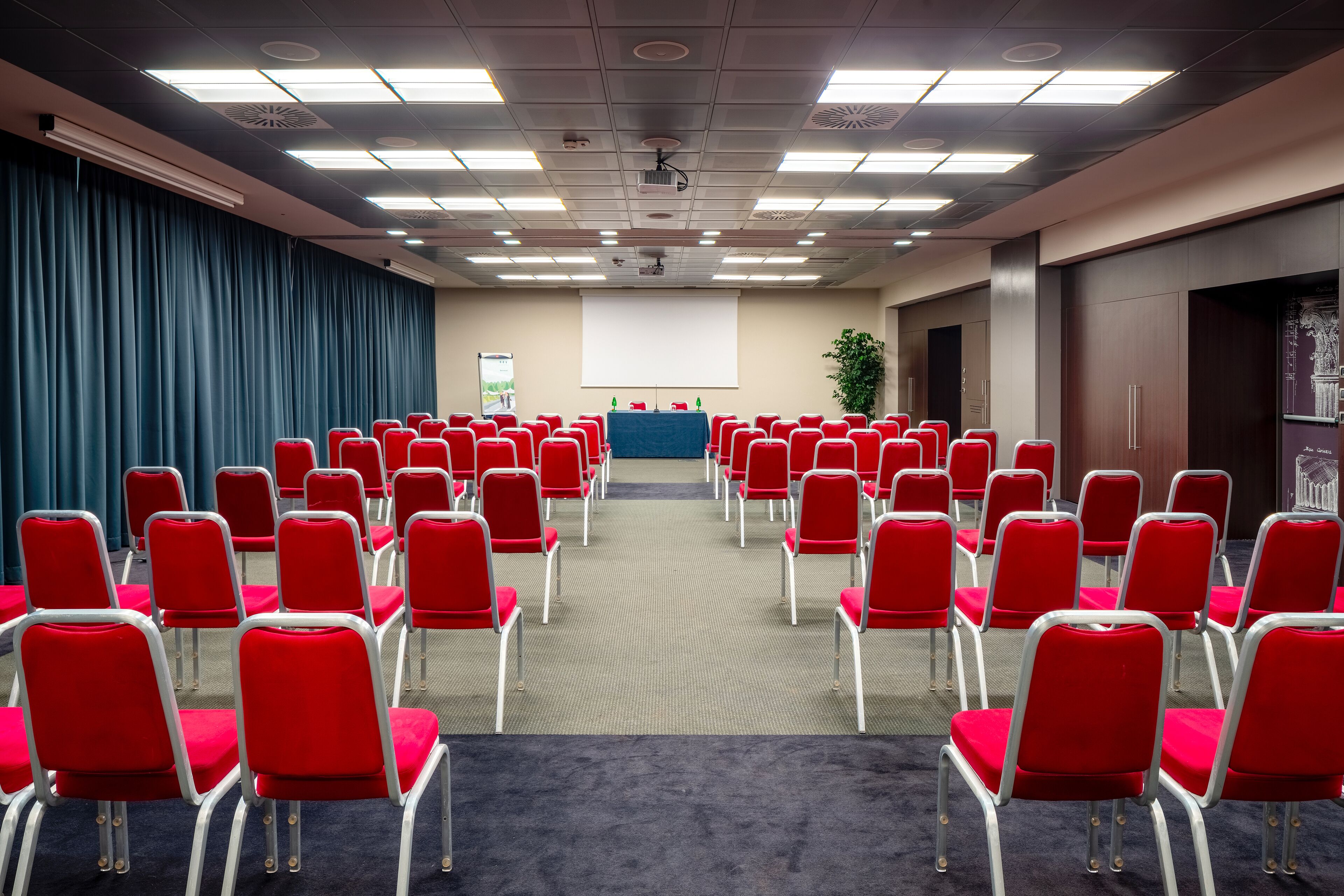 Uma sala de conferências com fileiras de cadeiras vermelhas, uma tela de projetor e um espaço para apresentações.