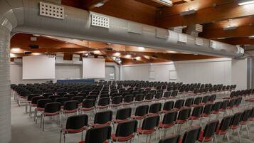 Uma sala de conferências com fileiras de cadeiras, uma tela de projeção e dutos de ventilação.