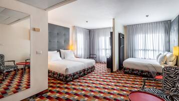 Quarto de hotel com uma cama grande, uma cama menor, uma escrivaninha, uma cadeira, uma televisão e um piso estampado.