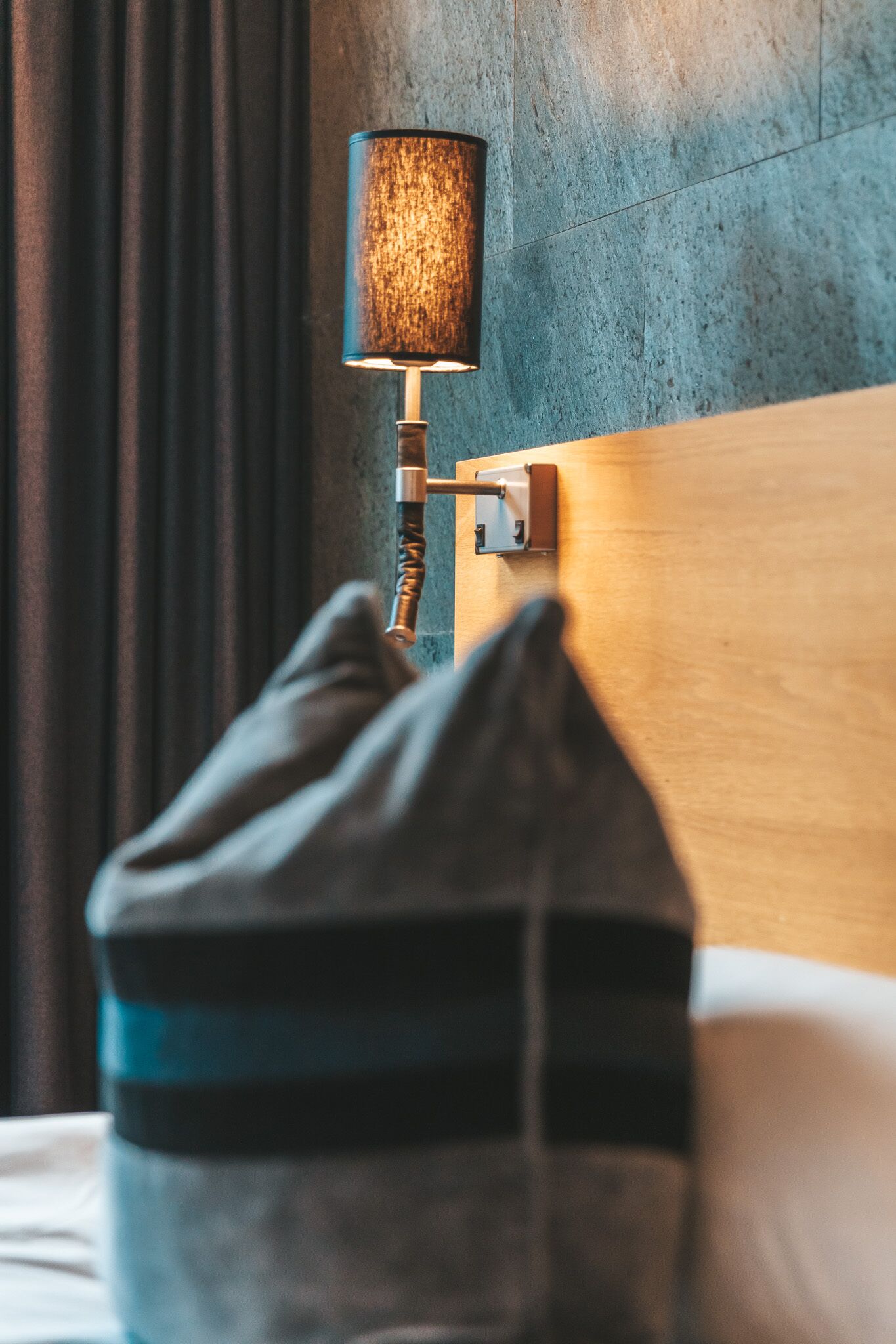 Quarto de hotel moderno com cabeceira de madeira, luminária fixada na parede e travesseiro de cor escura na cama.