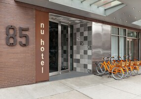 Entrada de hotel com um grande letreiro com o número 85, porta de vidro e uma fileira de bicicletas laranja estacionadas na frente.