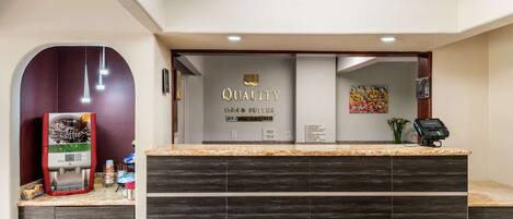 Uma área de recepção de hotel com uma máquina de café, uma tela digital e uma placa com a inscrição 'QUALITY INN & SUITES BY-CHOICE HOTELS'.