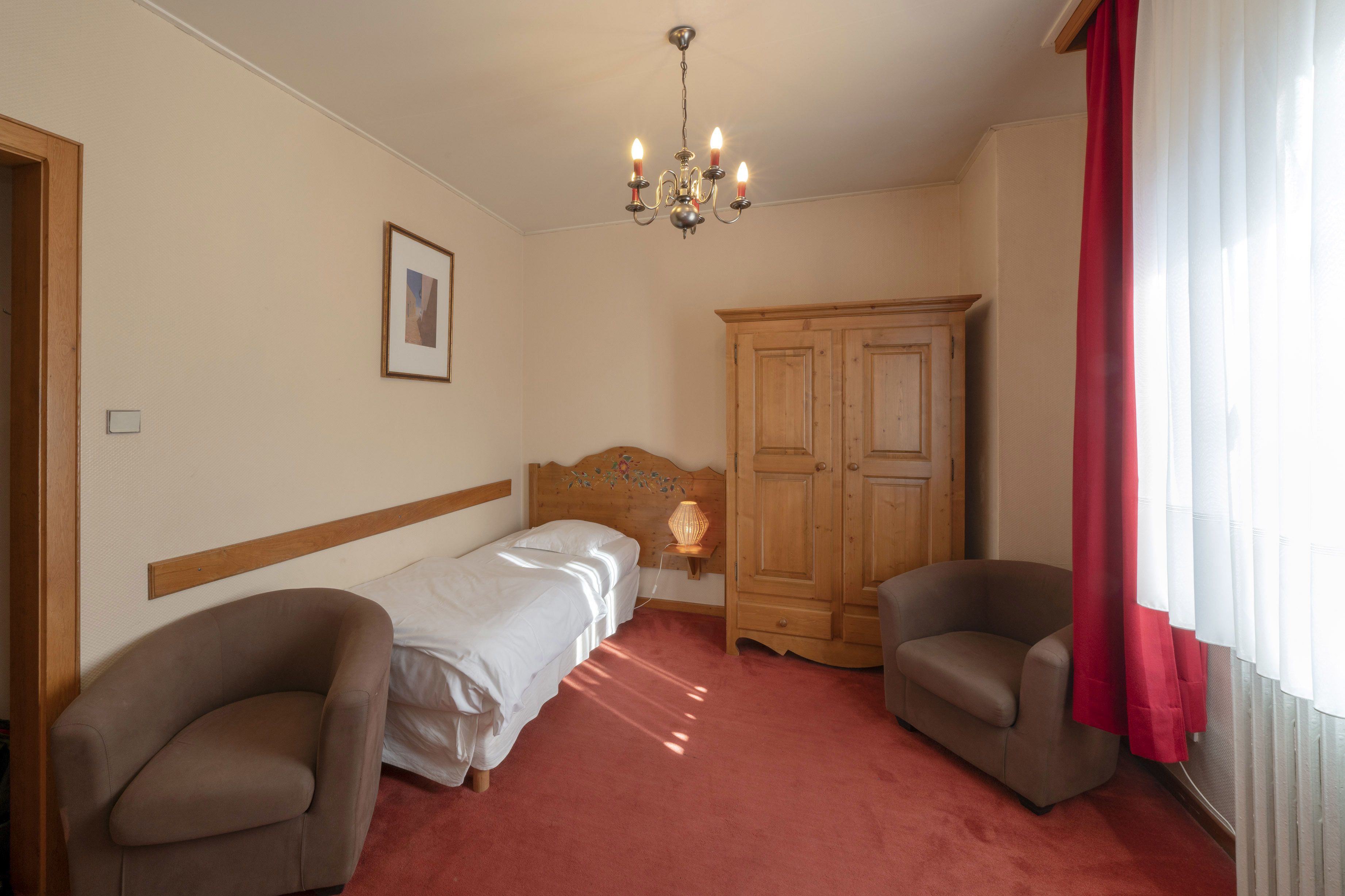Quarto de hotel com uma cama de solteiro, duas poltronas, um guarda-roupa de madeira, um candelabro e um quadro na parede.