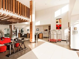 Um hall amplo com teto alto, balcão de recepção, área de refeições com detalhes em vermelho e um grande vaso de plantas.