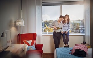 Quarto de hotel com uma poltrona vermelha, uma cama com colcha azul, uma escrivaninha com um laptop e uma janela com vista para a cidade.