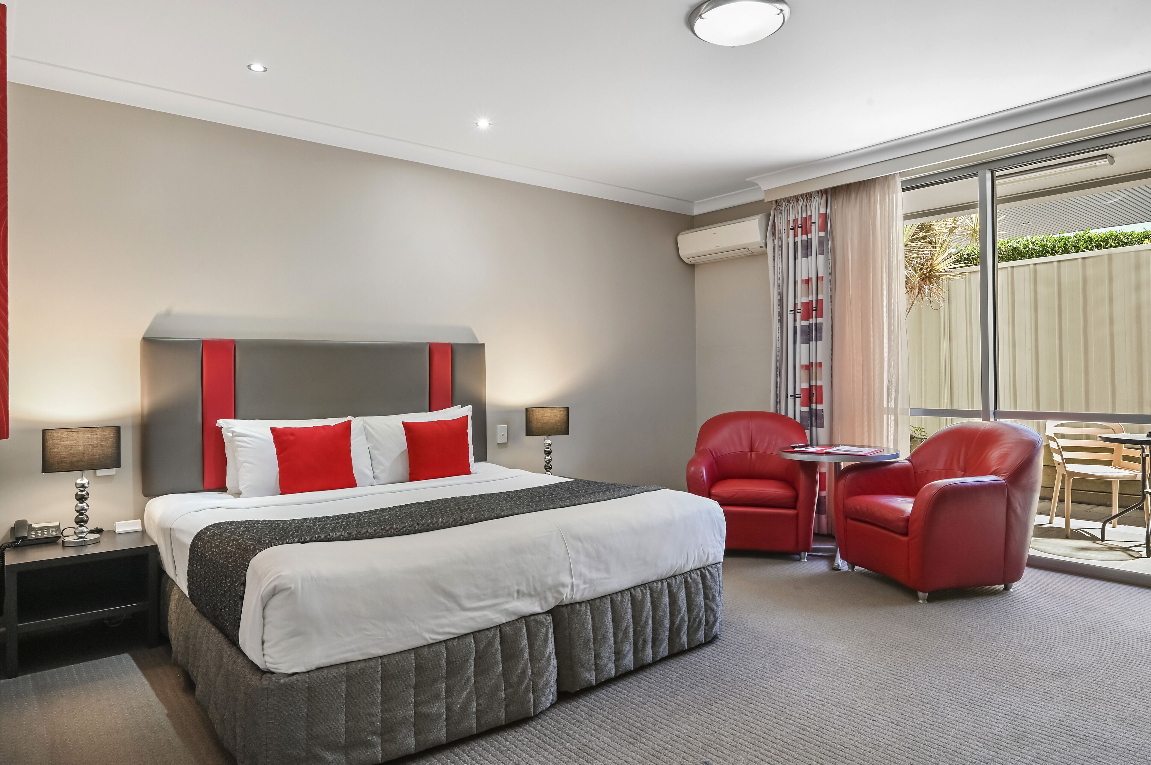 Quarto de hotel com uma cama grande, cadeiras vermelhas, uma mesa pequena e vista para o exterior.
