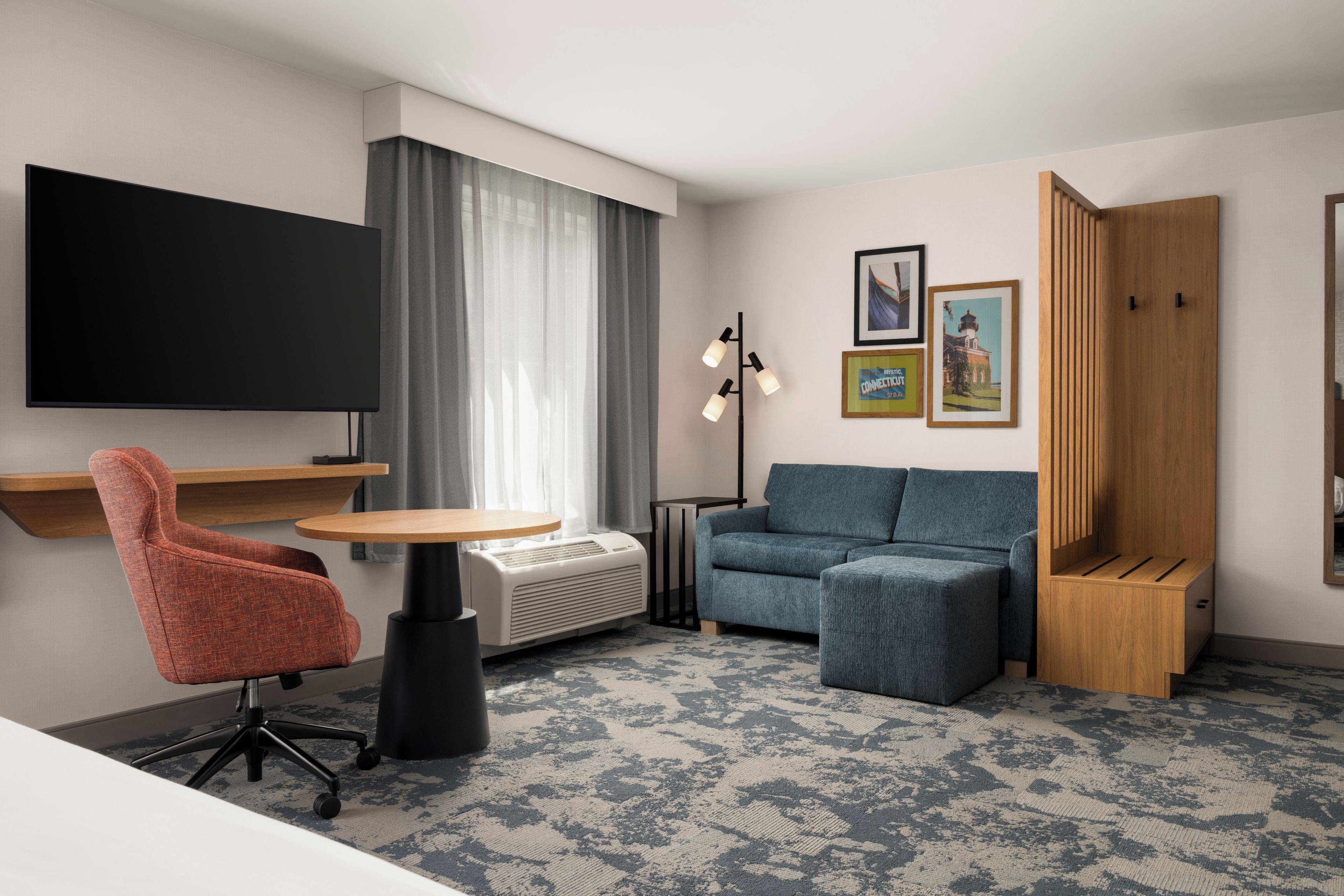 Quarto de hotel moderno com televisão de tela plana, sofá, cadeira, mesa pequena e luminária de chão.