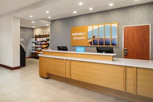 Bancada de recepção de hotel com uma placa 'Welcome to Hampton', um computador e uma vitrine de lanches.