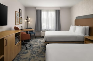Quarto de hotel com cama, escrivaninha, cadeira, televisão e janela com cortinas.