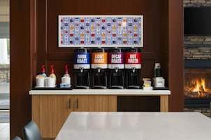 Estação de café com quatro dispensadores identificados como TORRA ESCURA, TORRA CLARA, DESCAFEINADO e ÁGUA QUENTE, e um expositor de xícaras de café na parede acima.