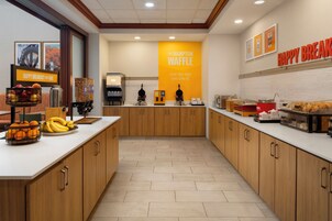 Cozinha moderna com ilha central, equipada com máquina de waffles, dispensador de suco e diversos alimentos.