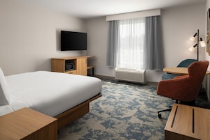 Quarto de hotel com cama, escrivaninha, cadeira, televisão, aparelho de ar condicionado e janela com cortinas.