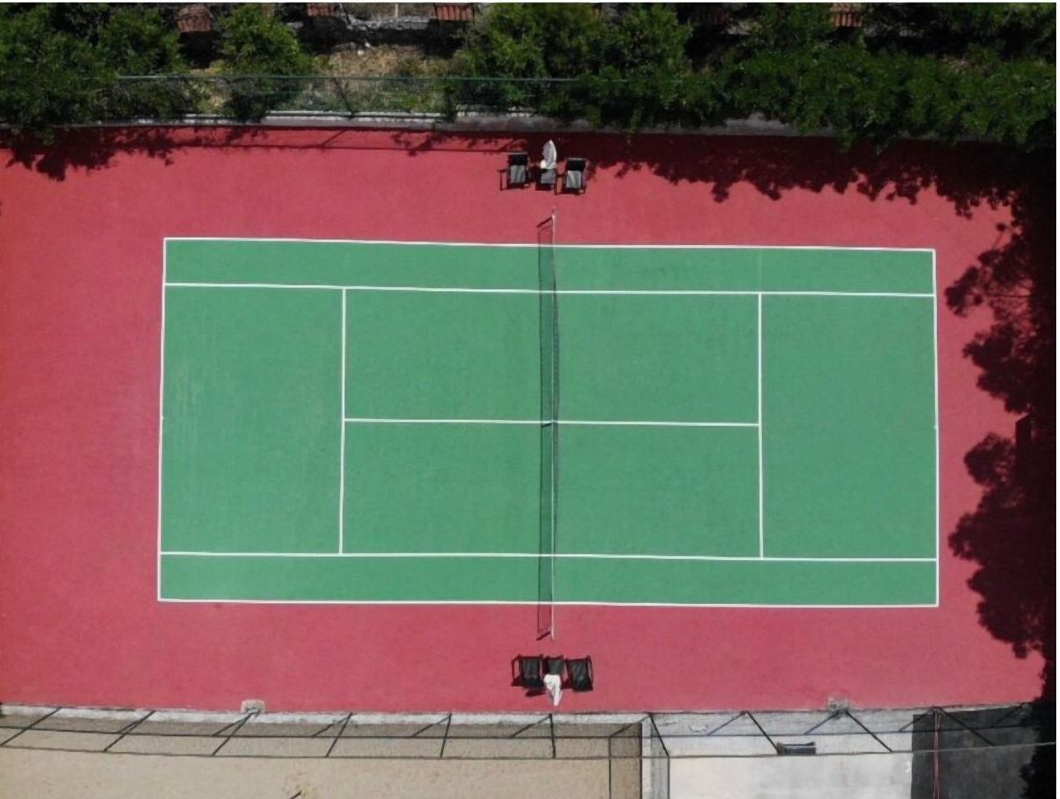 Vista aérea de uma quadra de tênis com a rede no centro, cercada por uma superfície vermelha e áreas verdes.