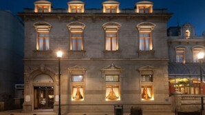 Um edifício histórico com fachada simétrica, grandes janelas com cortinas e uma entrada central com porta e degraus.